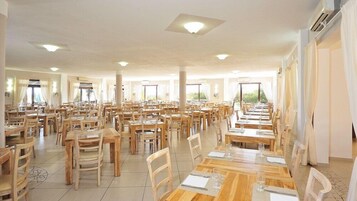 Uma ampla área de refeições com várias mesas e cadeiras de madeira, grandes janelas que permitem a entrada de luz natural e aparelhos de ar condicionado nas paredes.