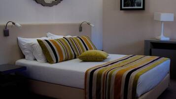 Quarto de hotel com cama, almofadas, mesa de cabeceira com abajur, espelho fixado na parede e quadro emoldurado.