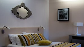 Quarto de hotel com cama, almofadas, mesa de cabeceira com abajur, espelho fixado na parede e quadro emoldurado.