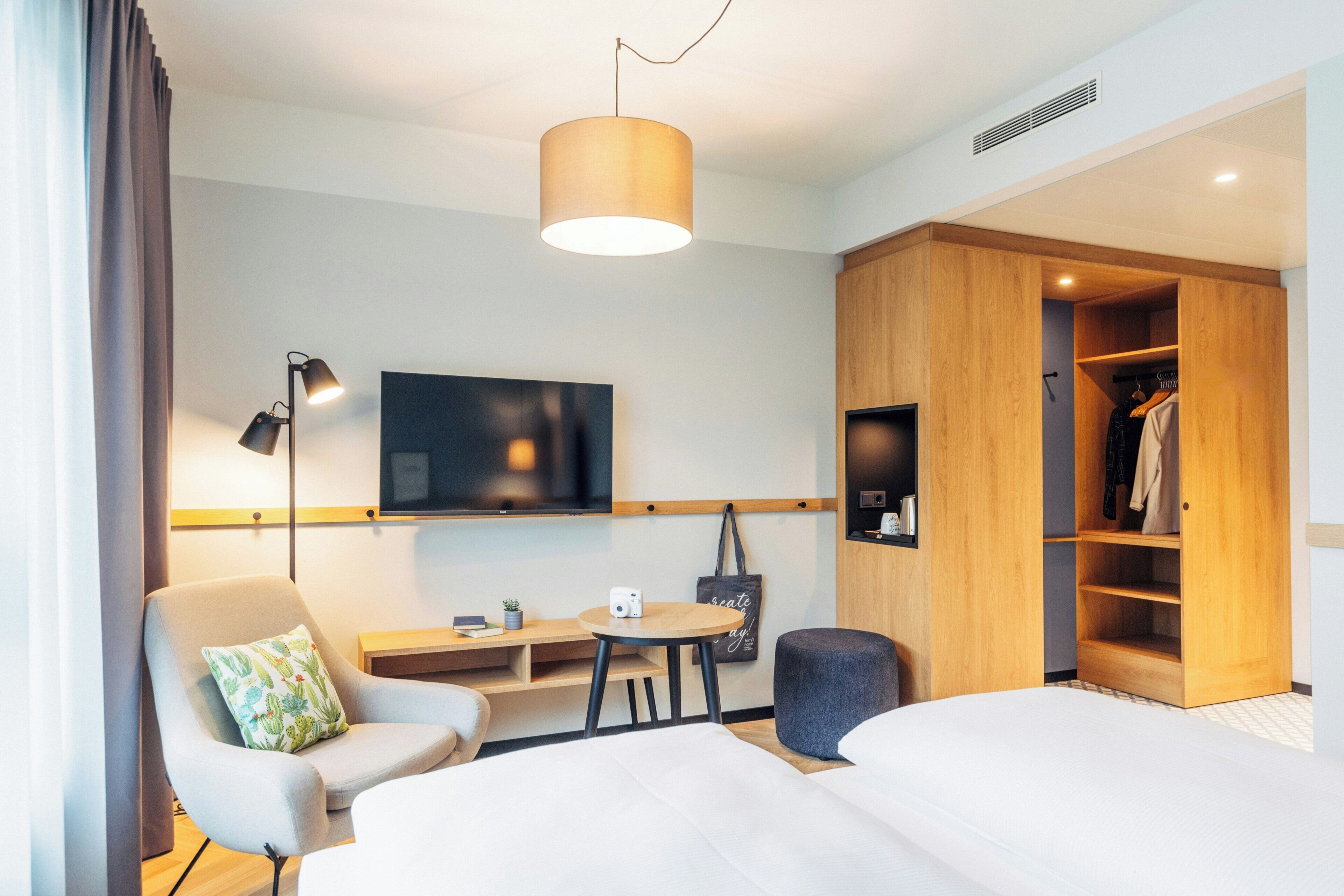 Quarto de hotel moderno com cama, escrivaninha, cadeira, televisão, guarda-roupa e cabideiro.