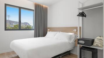 Quarto de hotel com uma cama, um pequeno frigorífico, uma televisão, uma janela com cortinas e vista para o exterior.