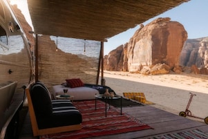 Uma casa de rodas com área de estar coberta, um sofá, uma pequena mesa e vista para uma paisagem desértica com formações rochosas.