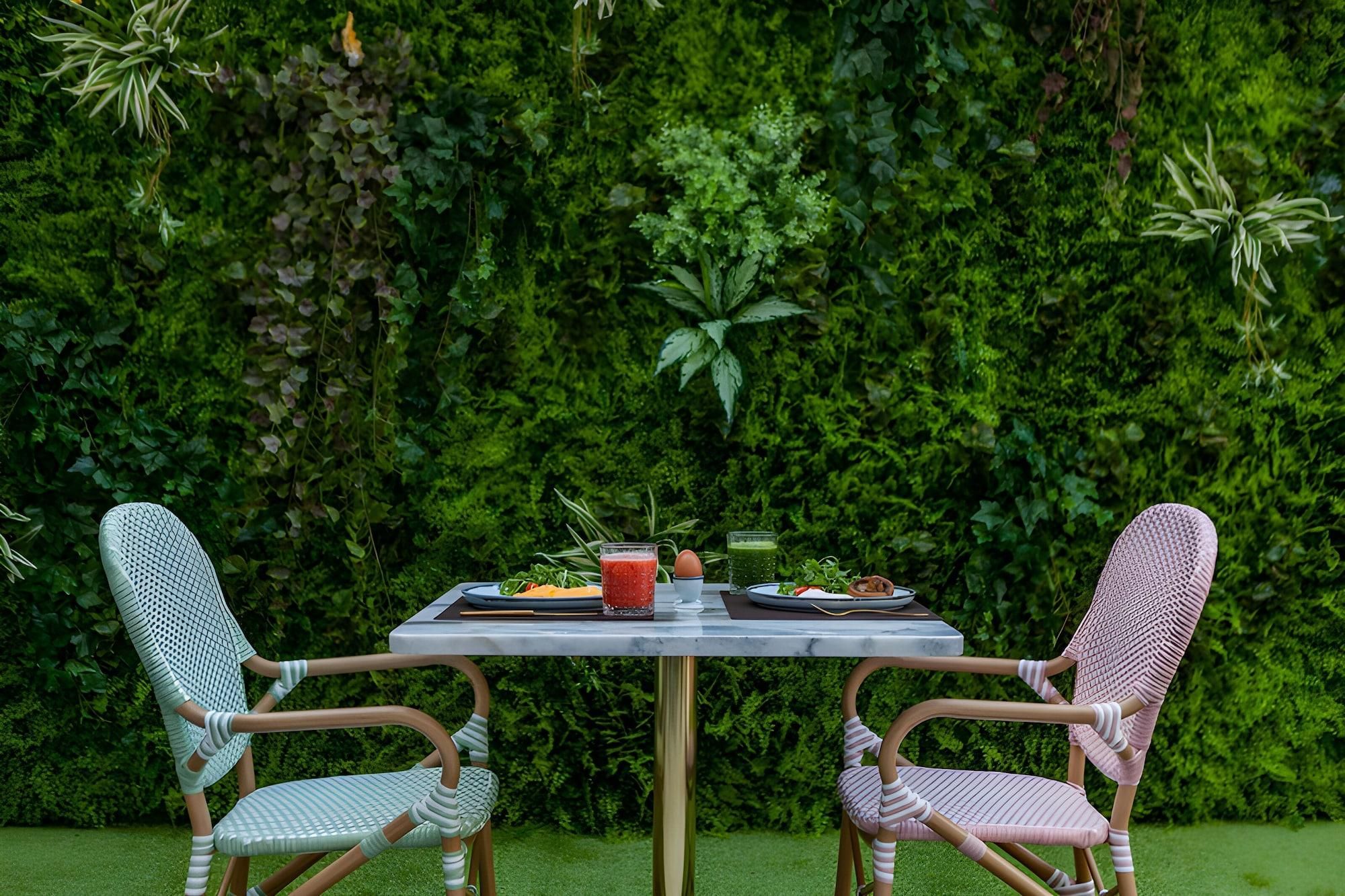 Uma área de refeições ao ar livre com uma mesa e duas cadeiras, rodeada por uma sebe verdejante.