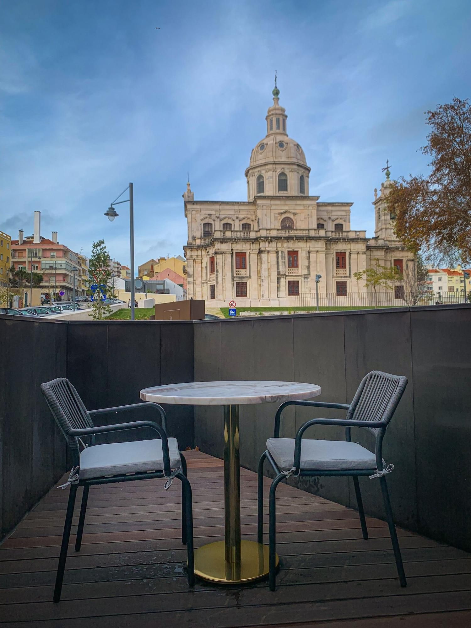 Uma área de estar ao ar livre com uma mesa redonda e duas cadeiras, um edifício grande com uma cúpula ao fundo e um céu limpo.
