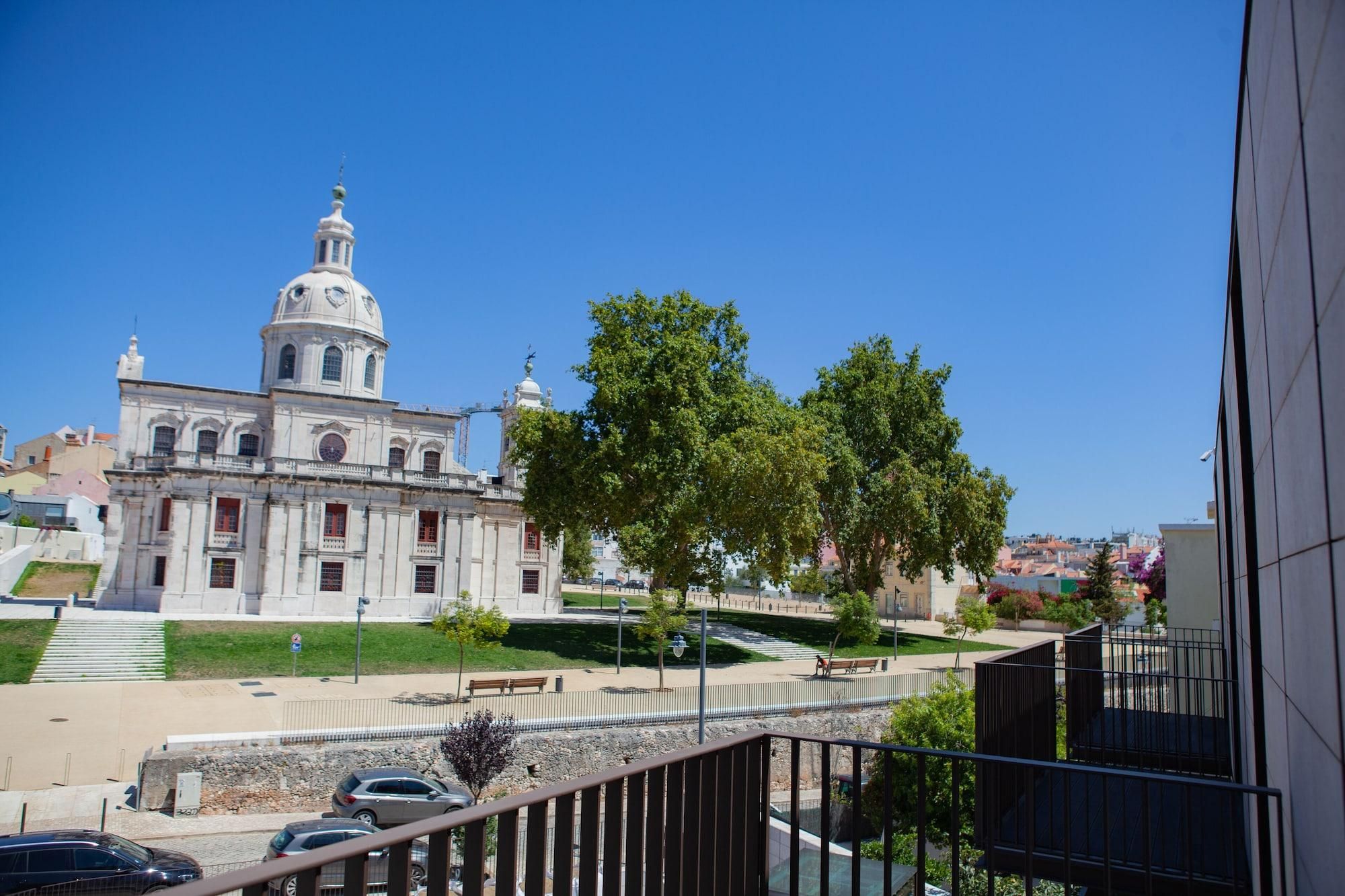 Um edifício histórico com cúpula e colunas, uma praça pavimentada com árvores, e um edifício moderno ao primeiro plano.