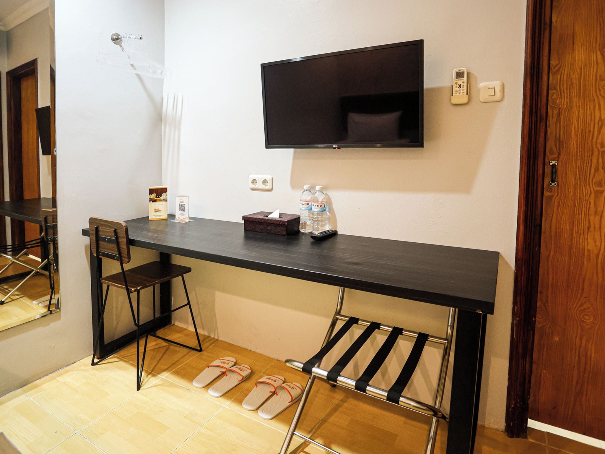 Quarto com escrivaninha, cadeira, televisão na parede, mesinha com caixa de lenços e garrafas de água, sapateira e porta de madeira.