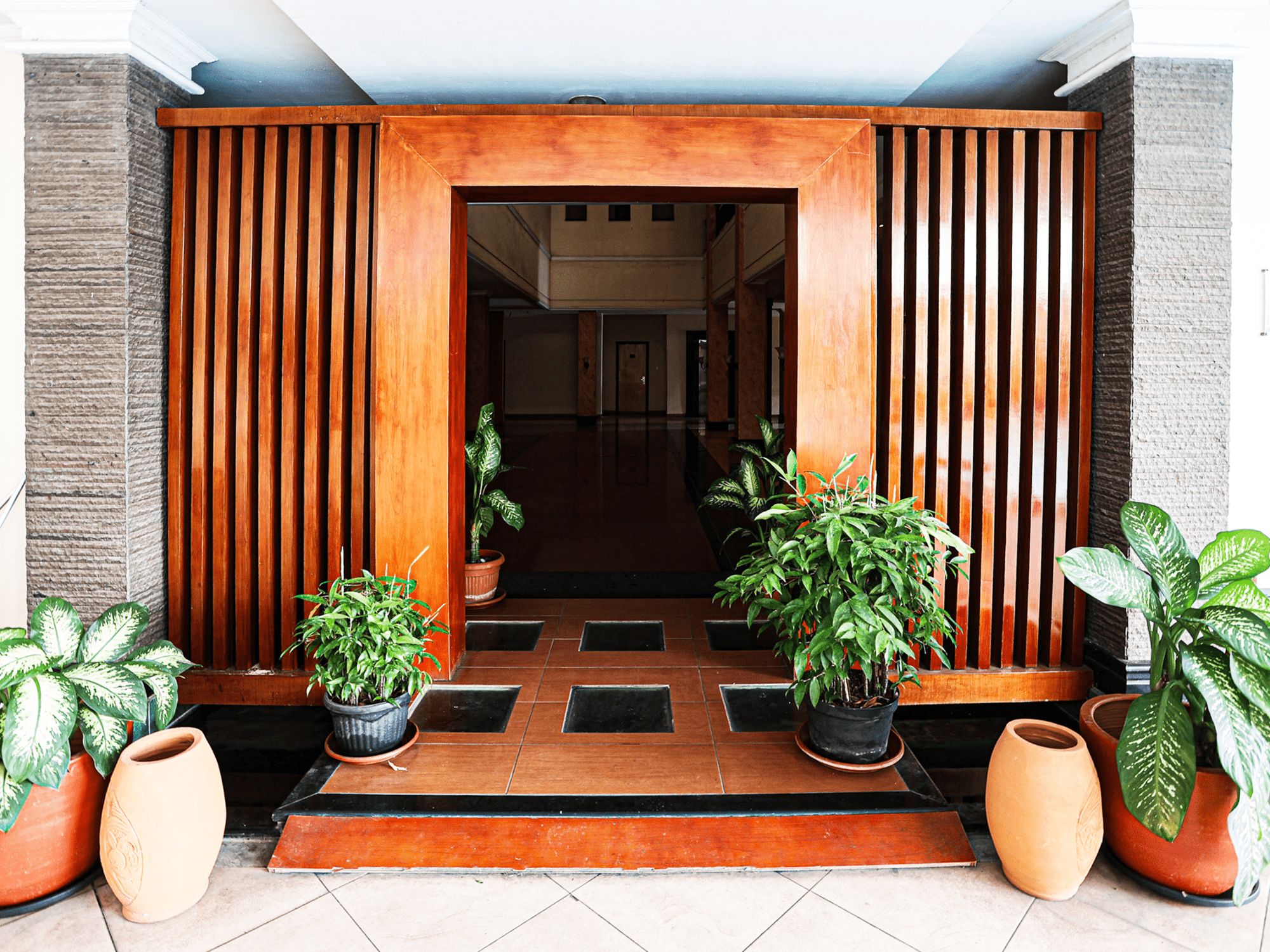 Entrada de hotel com fachada de madeira, plantas em vasos e piso de azulejos.
