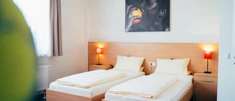 Quarto de hotel com duas camas de solteiro, mesa de cabeceira com abajur, obra de arte na parede e janela com cortinas.