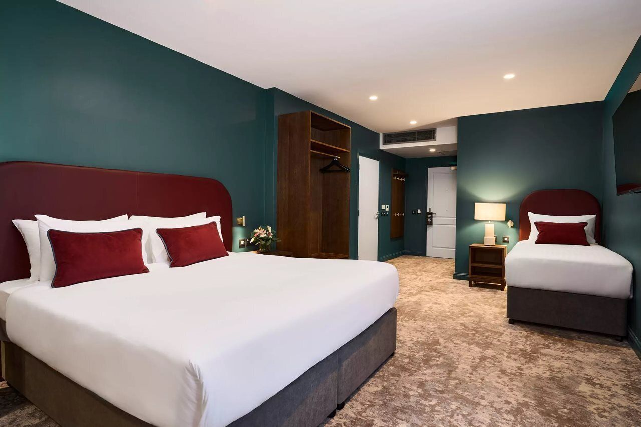 Quarto de hotel com uma cama grande, uma cama menor, um guarda-roupa, uma mesa de cabeceira e uma luminária.
