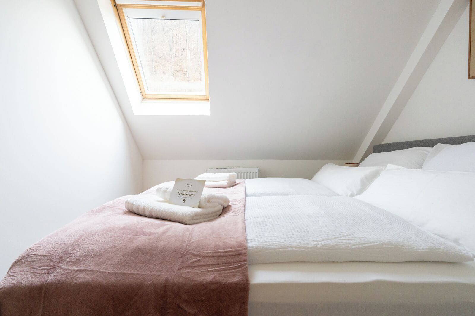 Quarto com teto inclinado, claraboia, cama com roupa de cama branca e placa indicando 10% de desconto no serviço de quarto.