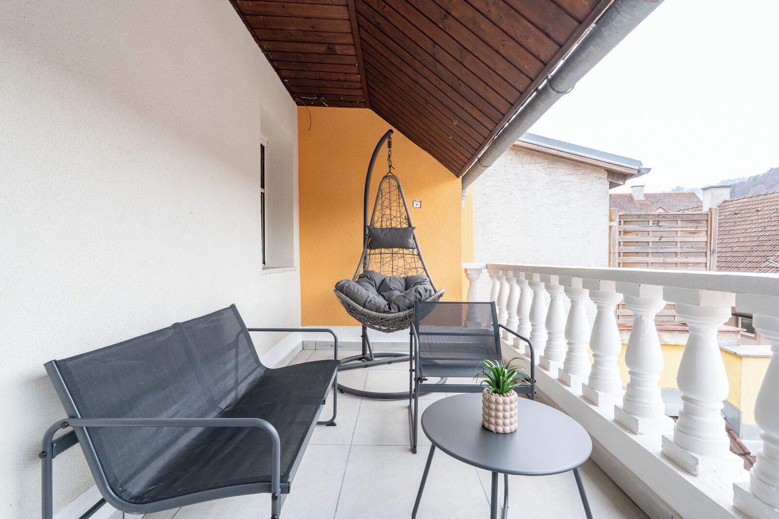 Varanda com sofá preto, cadeira suspensa e pequena mesa com uma planta.