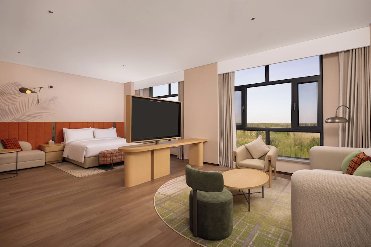 Quarto de hotel com cama, televisão, escrivaninha, sofá, poltrona e janelas amplas.