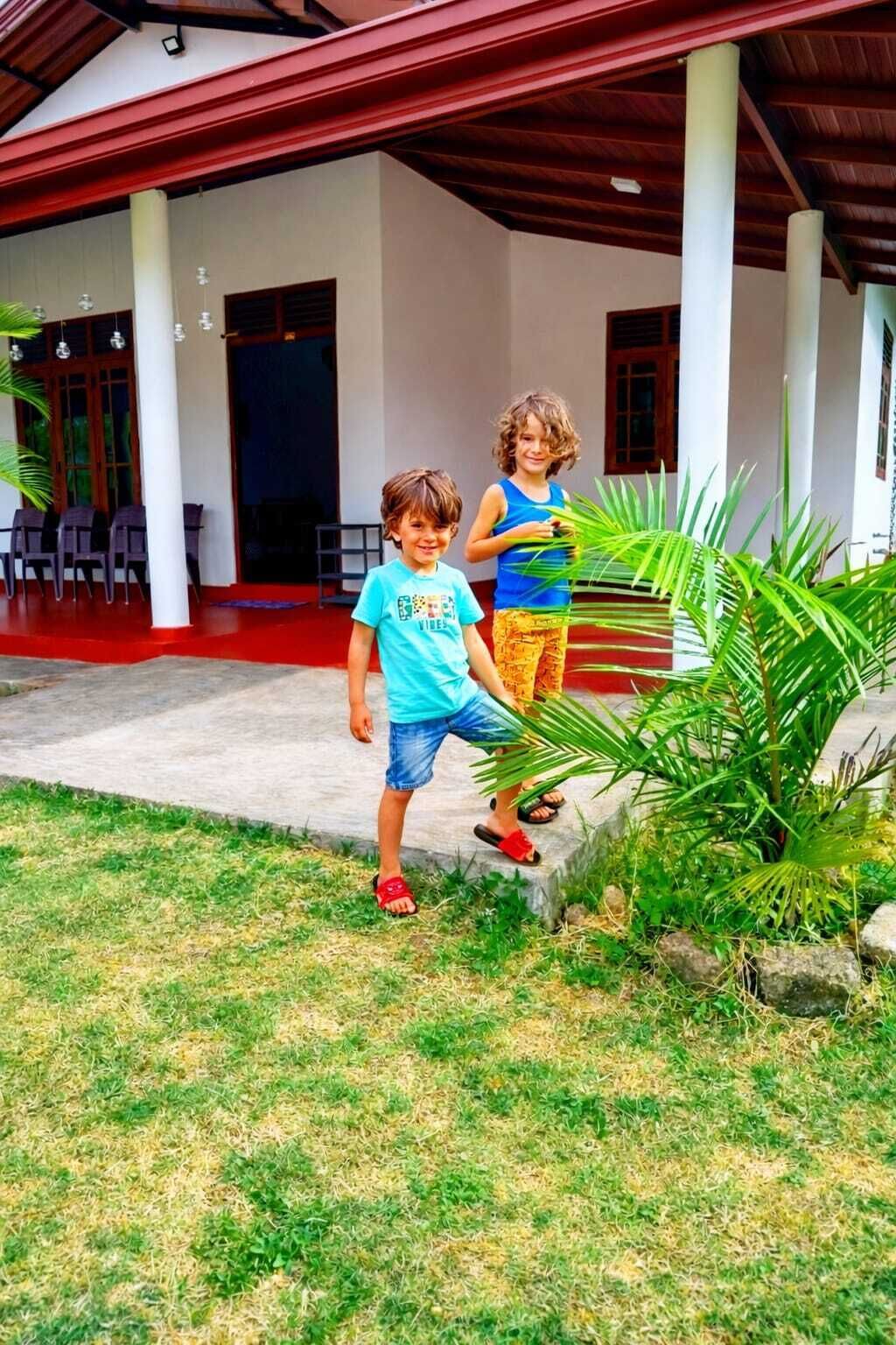 Duas crianças em um alpendre com telhado vermelho e colunas brancas, em meio à vegetação e um pequeno jardim.