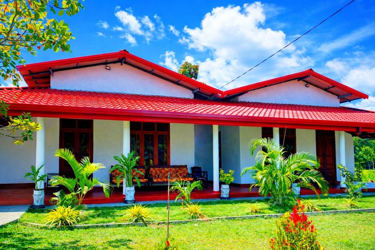 Uma casa de dois andares com telhado vermelho e paredes brancas, apresentando um alpendre coberto com cadeiras e plantas em vasos.