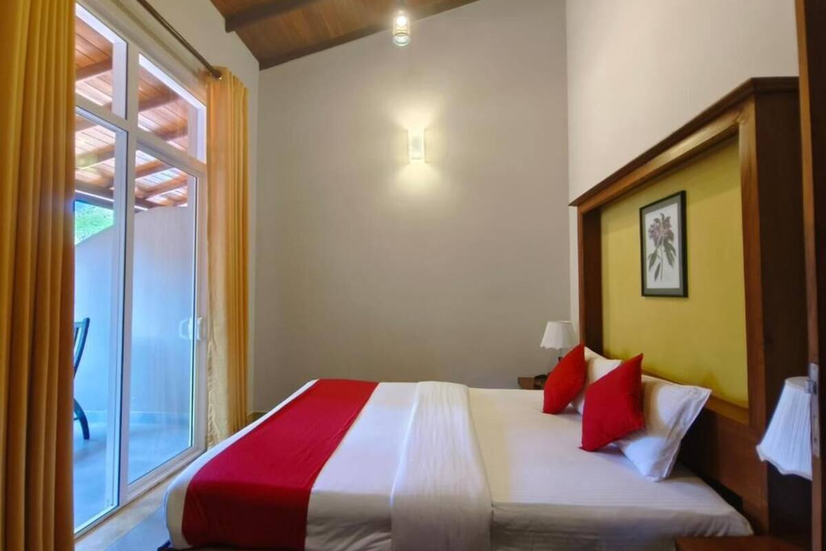 Quarto de hotel com cama, mesa de cabeceira, luminária e janela com cortinas.