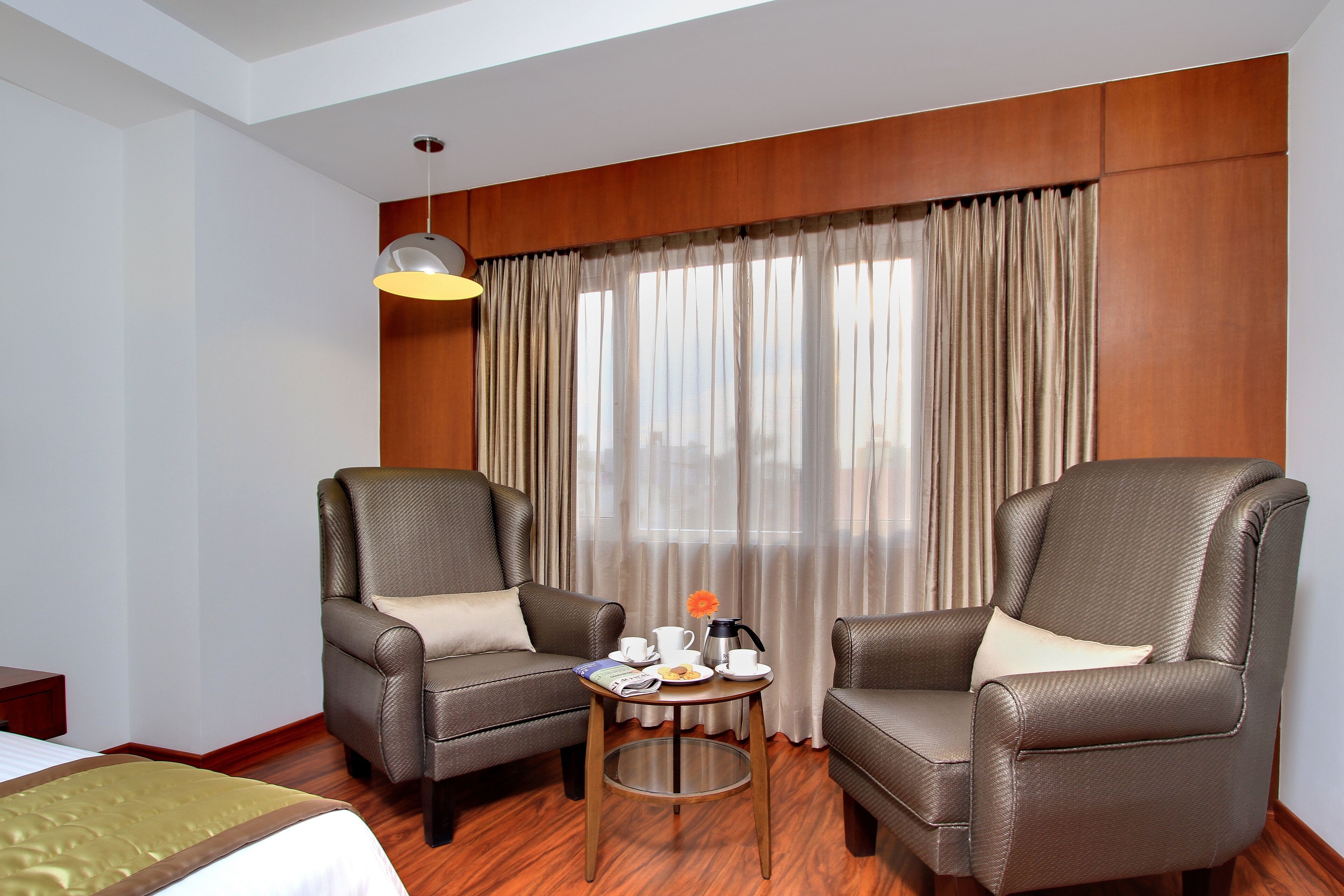 Quarto de hotel com duas poltronas de couro, uma mesa redonda pequena com uma cafeteira, xícaras e um livro, além de uma cama com cabeceira dourada.