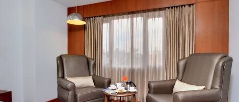 Quarto de hotel com duas poltronas de couro, uma mesa redonda pequena com uma cafeteira, xícaras e um livro, além de uma cama com cabeceira dourada.