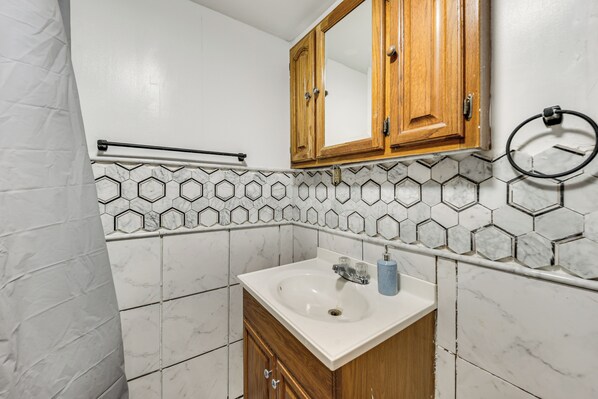 Casa de banho com lavatório branco, armários de madeira, espelho e painel de azulejos hexagonais.