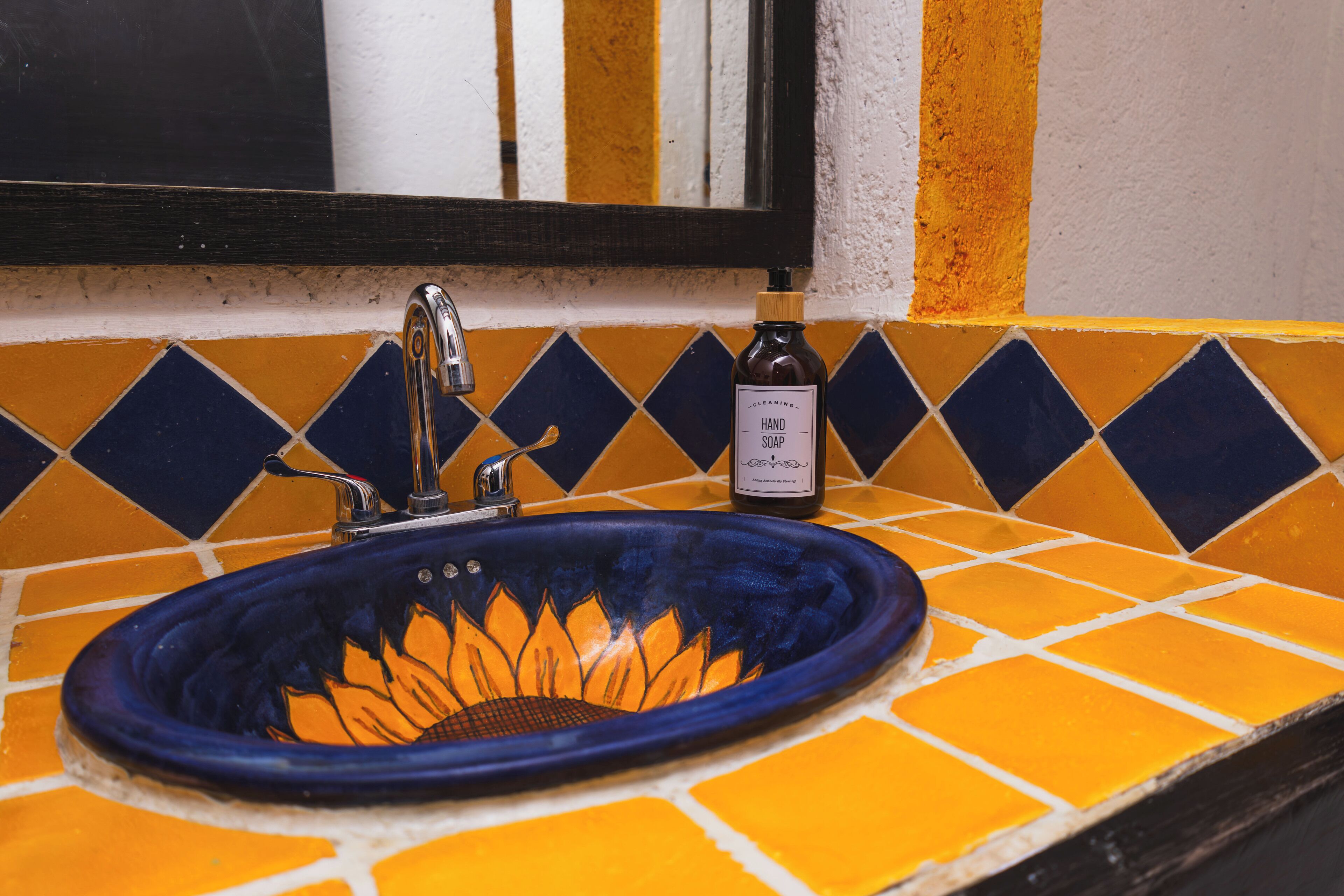 Uma pia de banheiro com cuba azul e design floral, um espelho acima e uma garrafa de sabonete líquido na parede revestida de azulejos.