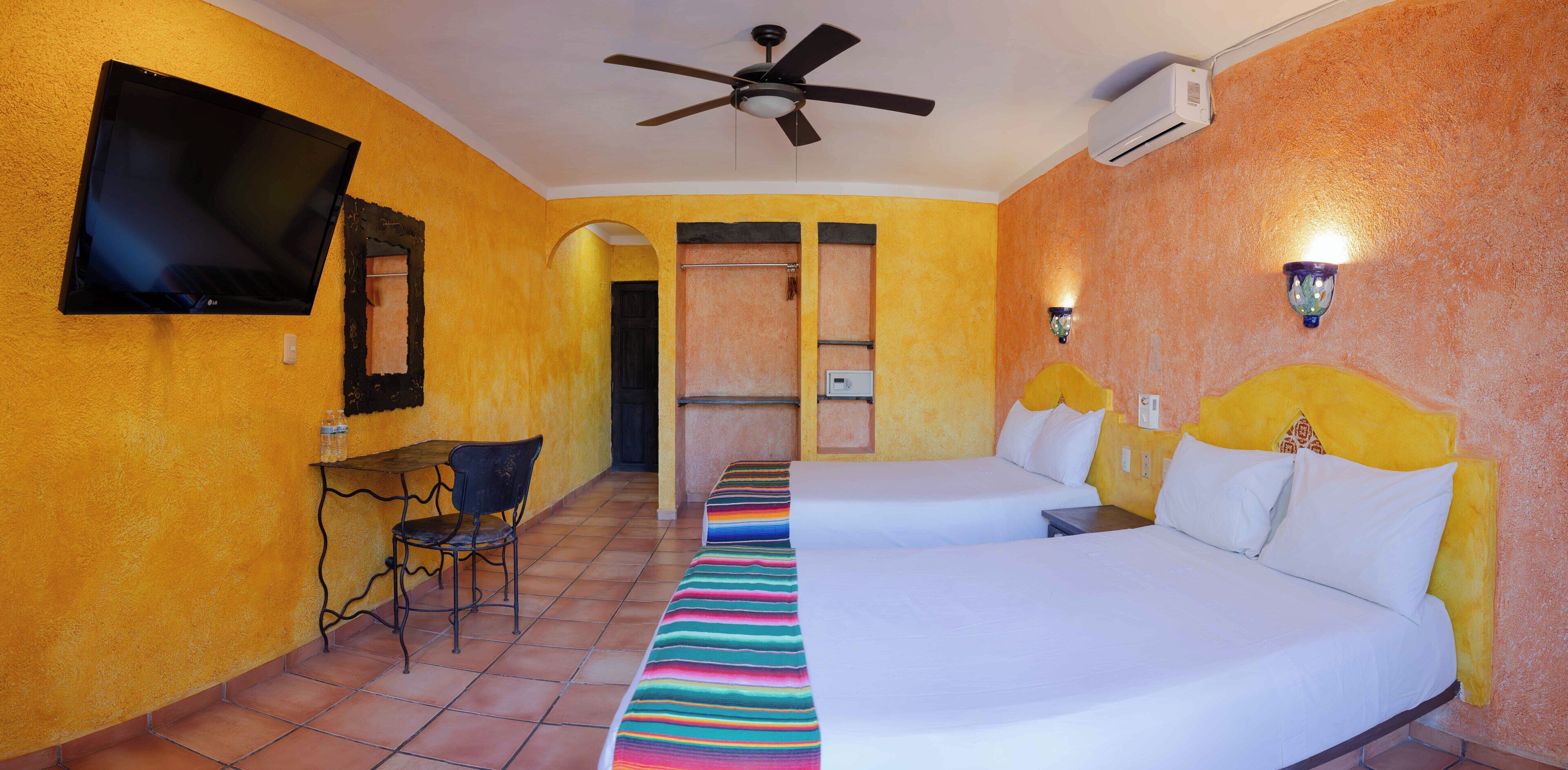 Quarto de hotel com duas camas, uma televisão, um ventilador de teto e um banheiro visível através de um arco.