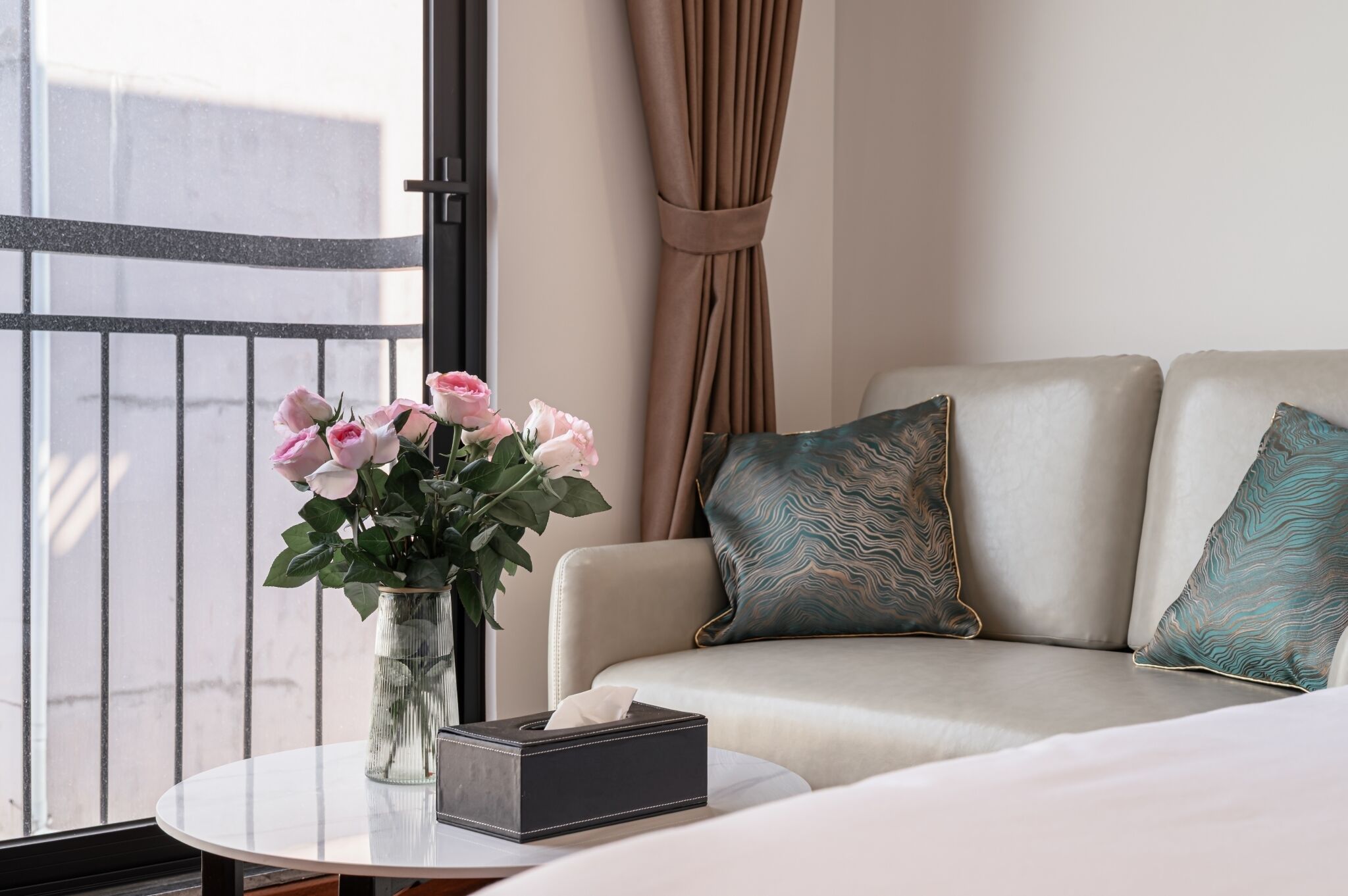 Quarto de hotel com sofá, vaso de flores, caixa de lenços e janela com cortina.