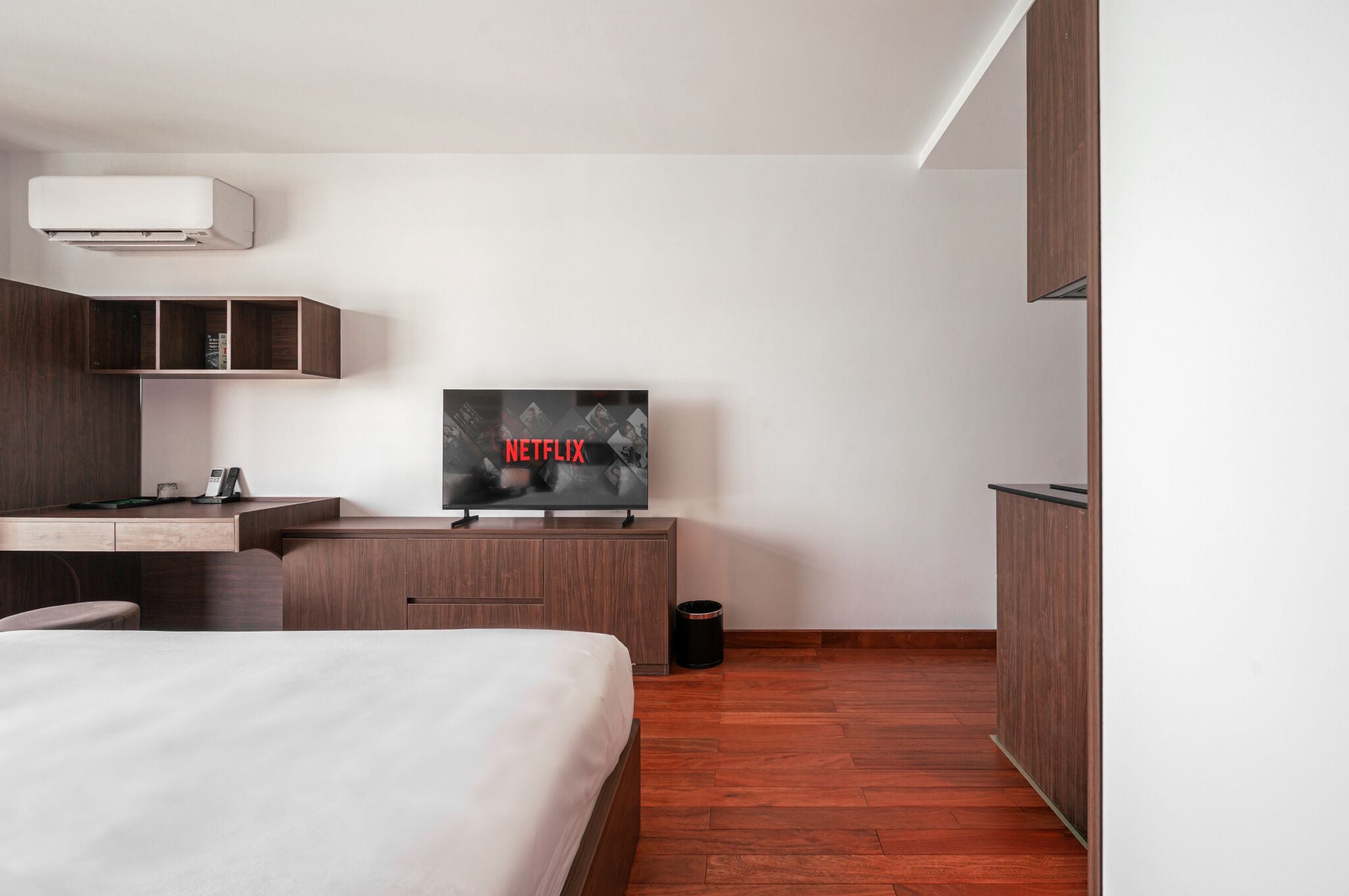 Quarto de hotel moderno com cama, escrivaninha, televisão exibindo o logotipo da Netflix, guarda-roupa e lixeira.