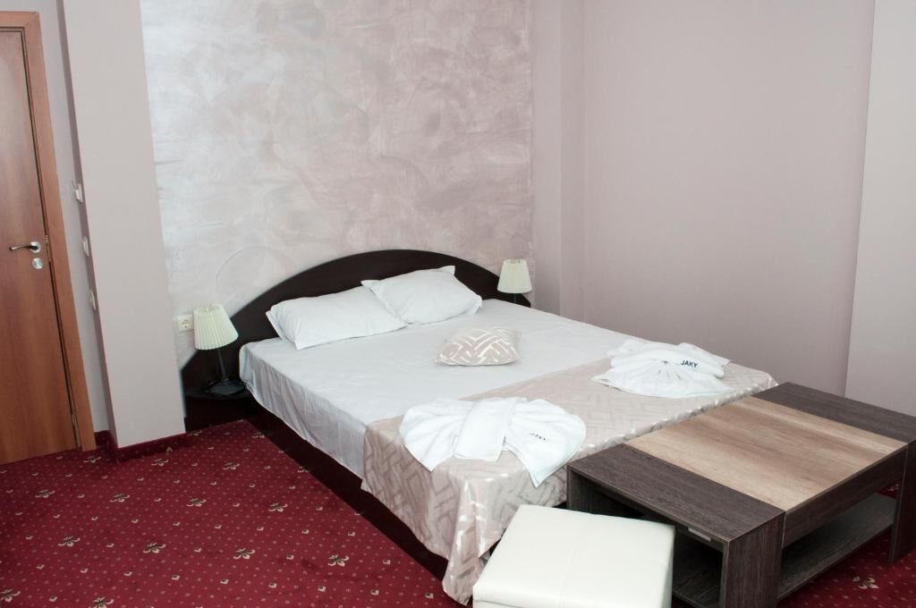 Quarto de hotel com cama, mesas de cabeceira, um criado-mudo de madeira e um tapete vermelho.