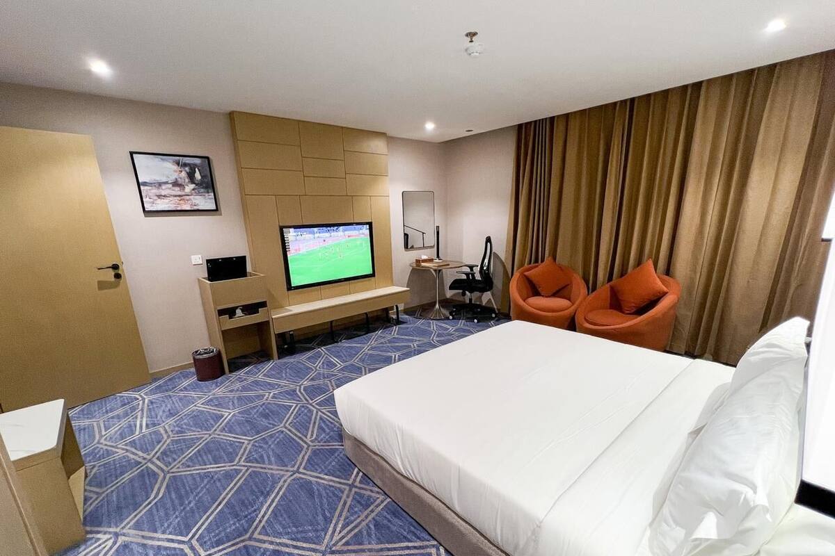Quarto de hotel com uma cama grande, televisão, escrivaninha e duas cadeiras laranja.