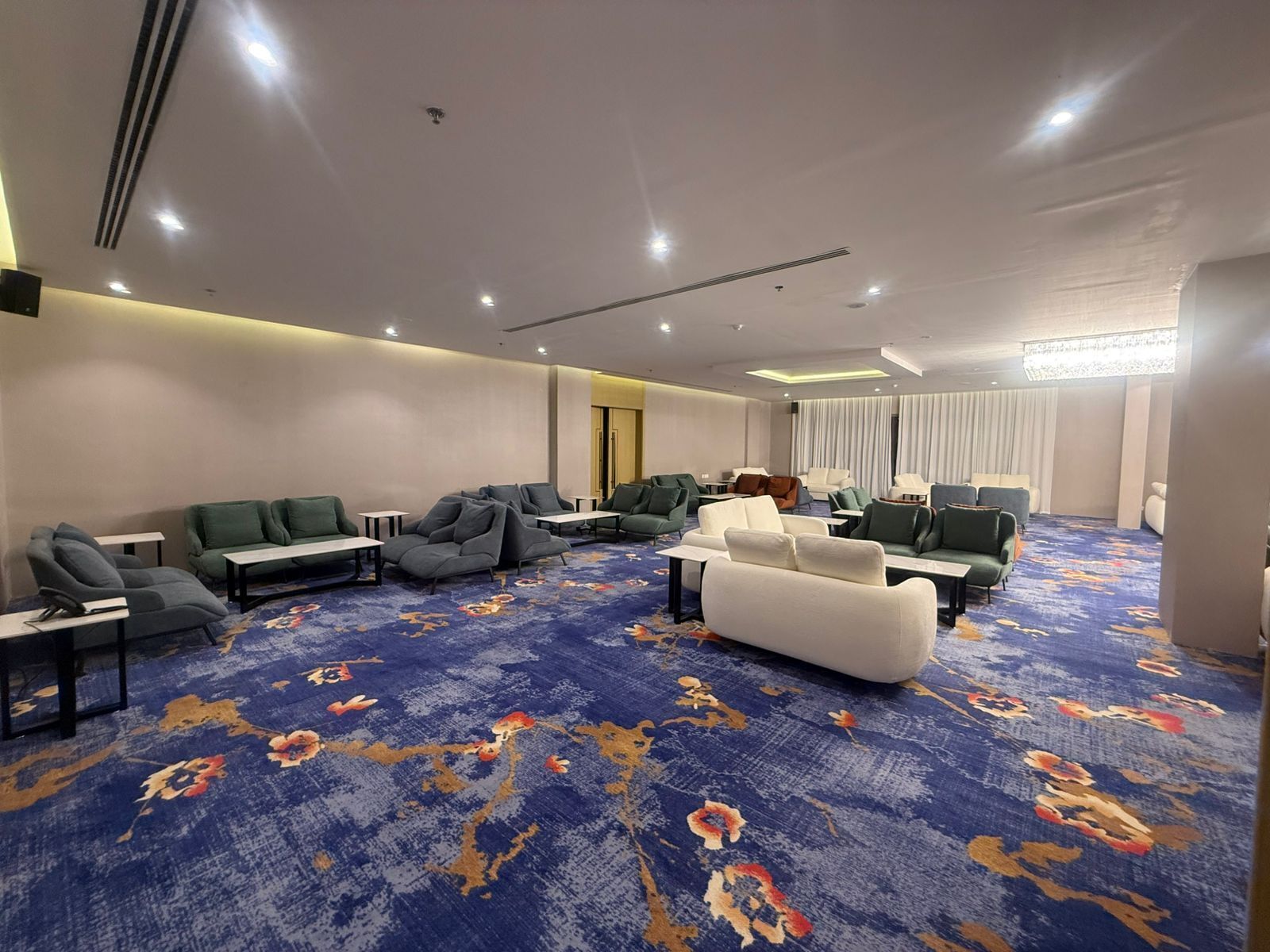 Um quarto espaçoso com carpete floral azul, várias áreas de estar com sofás e poltronas, e uma grande janela com cortinas.