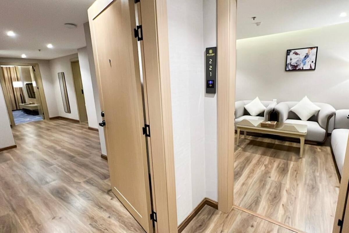 Quarto de hotel com piso de madeira, um sofá com almofadas, uma mesa pequena e um painel de controle fixado na parede.
