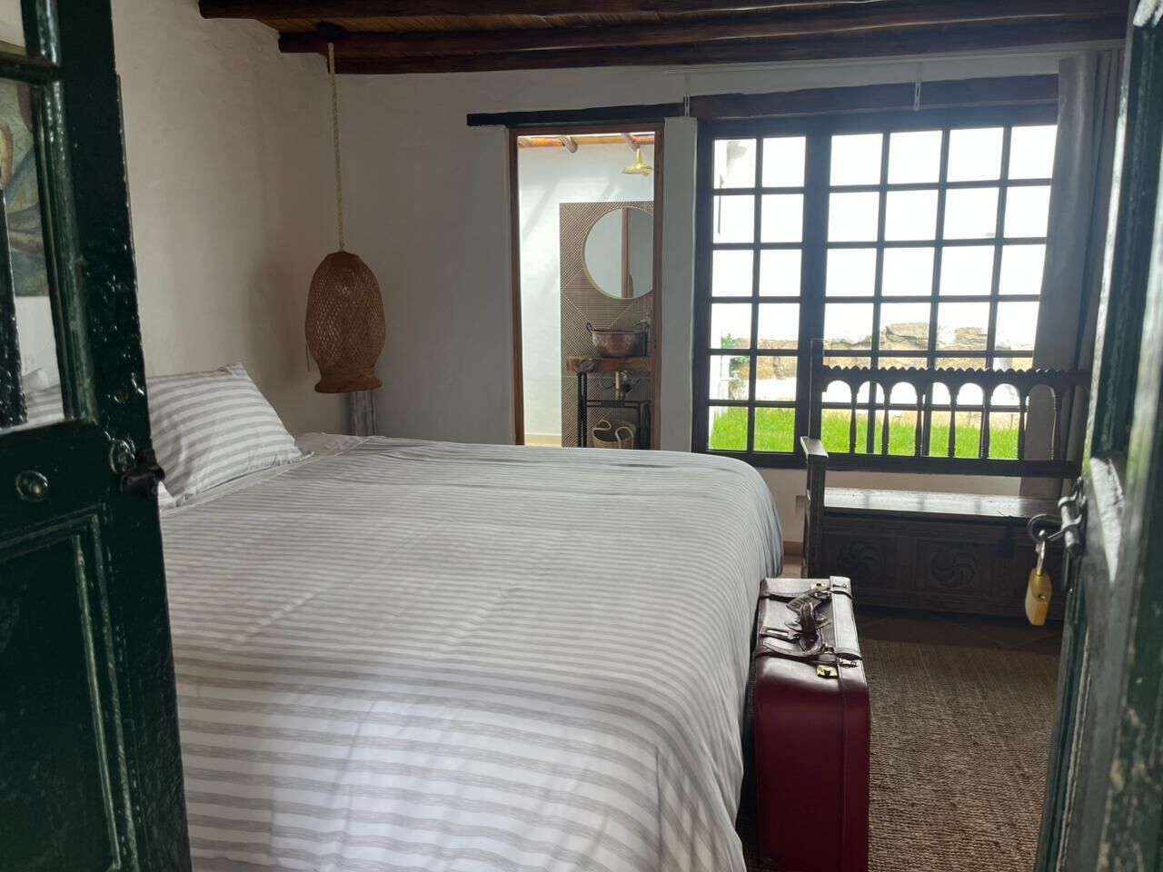 Quarto com cama, mala, baú de madeira, luminária pendente e vista para uma paisagem verde pela janela.