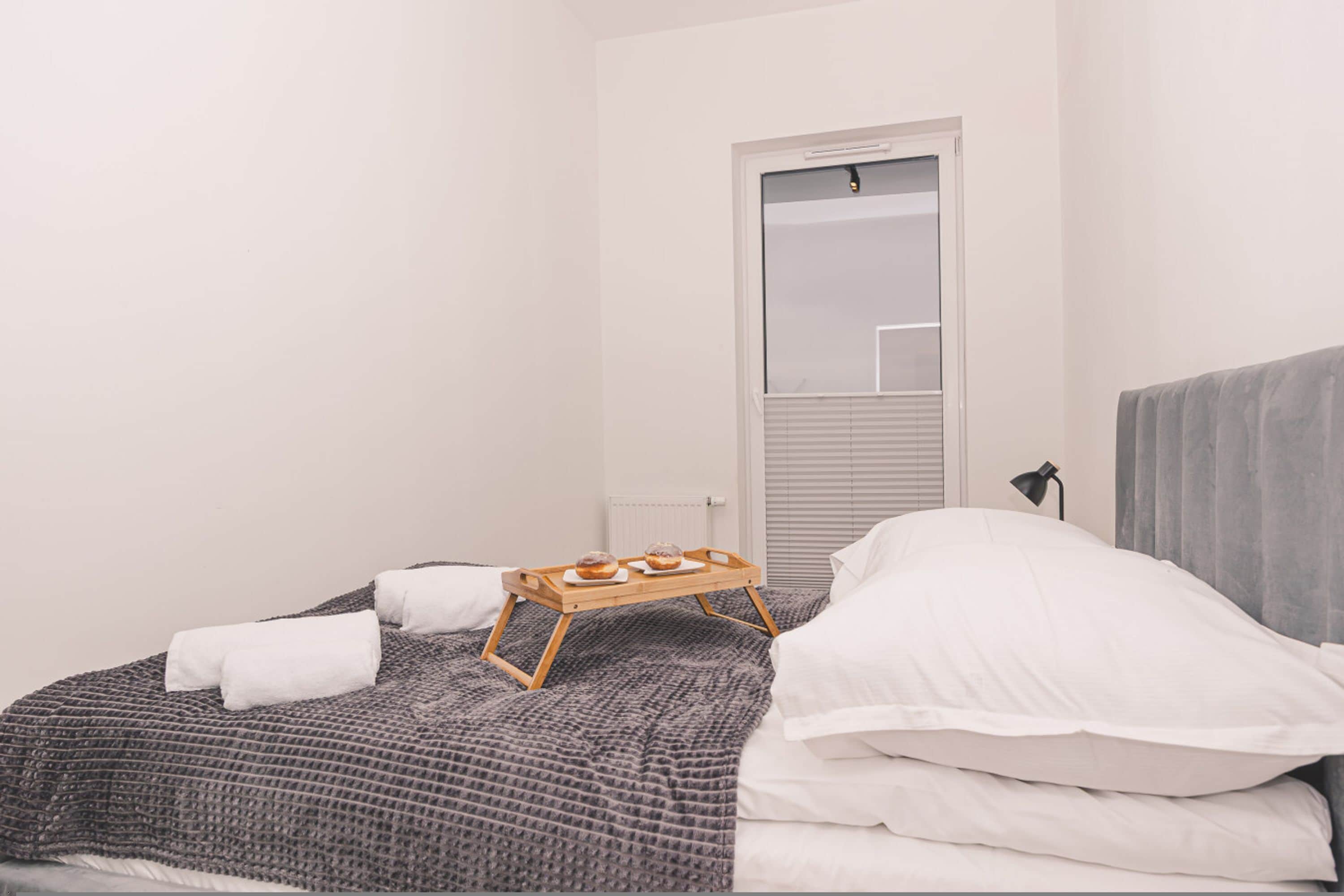Quarto com cama, mesa de cabeceira com bandeja de donuts, luminária e janela com persianas.