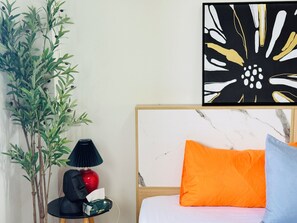 Quarto com cama, almofadas, luminária, planta e uma obra de arte na parede.
