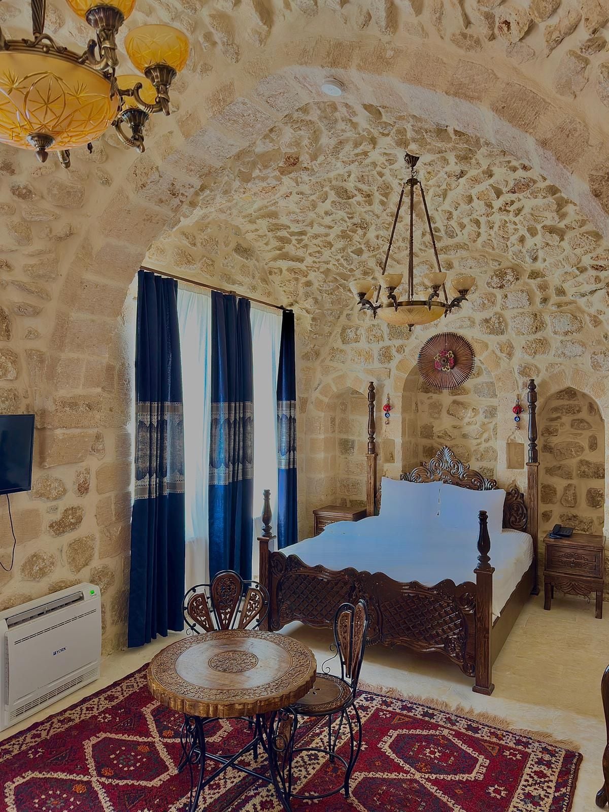 Quarto com parede de pedra, cama com cabeceira decorada, lustre, mesa redonda com cadeiras e tapete estampado.