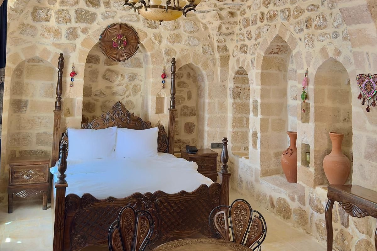 Quarto com parede de pedra, cama, lustre e mesa com cadeiras.