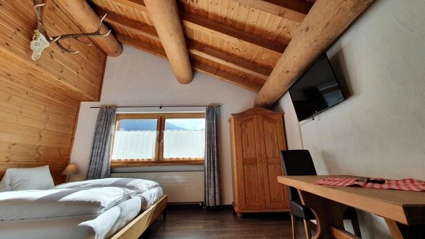 Quarto com vigas de madeira no teto, uma cama, uma escrivaninha e uma televisão fixada na parede.