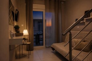 Uma sala de estar aconchegante com um sofá, uma luminária, uma mesinha com uma planta e vista para uma varanda externa através de uma porta de vidro.
