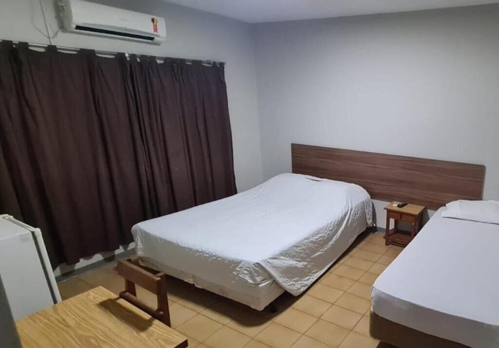 Quarto de hotel com uma cama de casal, mesa de cabeceira de madeira, uma mesinha e uma janela com cortinas.