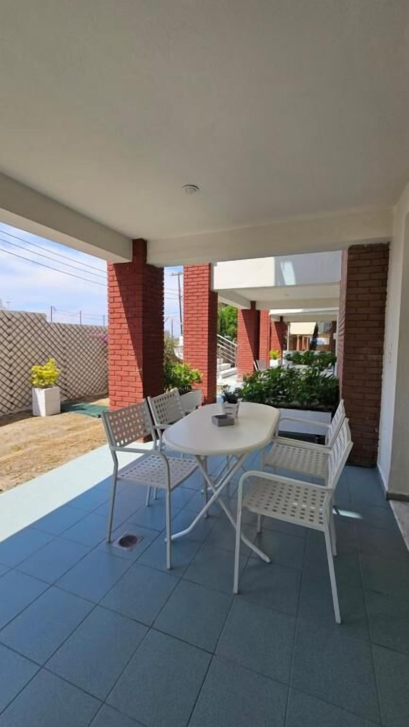 Uma varanda coberta com mesa e cadeiras, rodeada por pilares de tijolo e vista para um jardim.