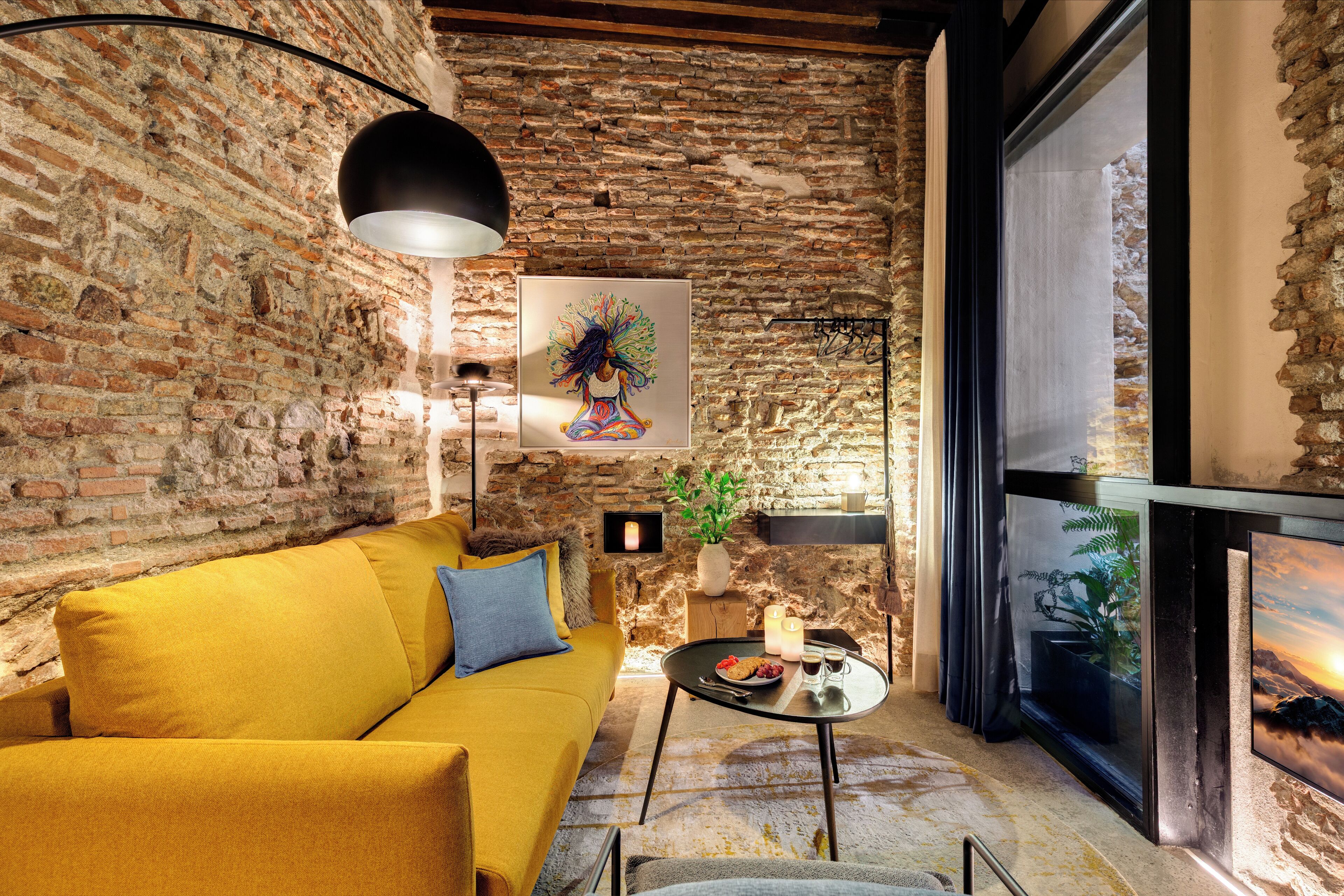 Um quarto acolhedor com um sofá amarelo, uma mesa redonda com velas, um quadro na parede e uma lareira com uma planta.