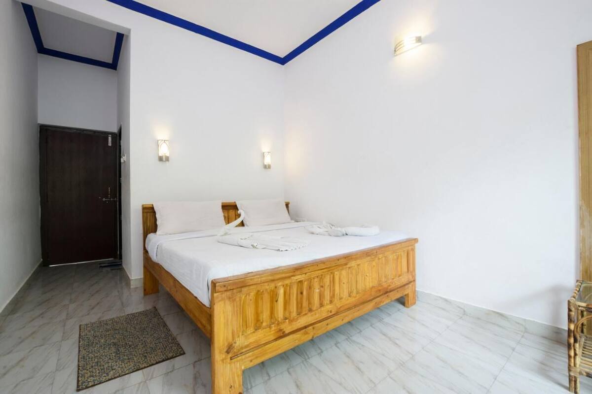 Quarto com cama de madeira, roupa de cama branca, porta escura e duas luzes fixadas na parede.