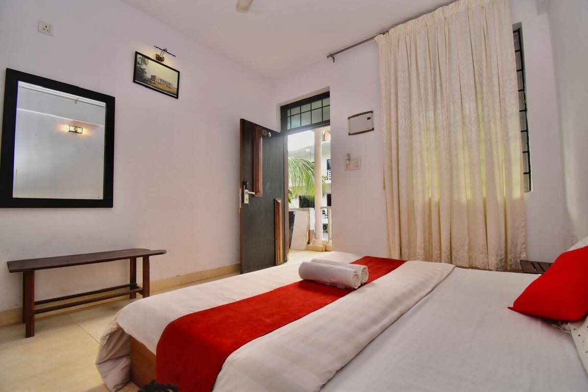 Quarto de hotel com uma cama, um banco, uma porta e uma janela com cortinas.