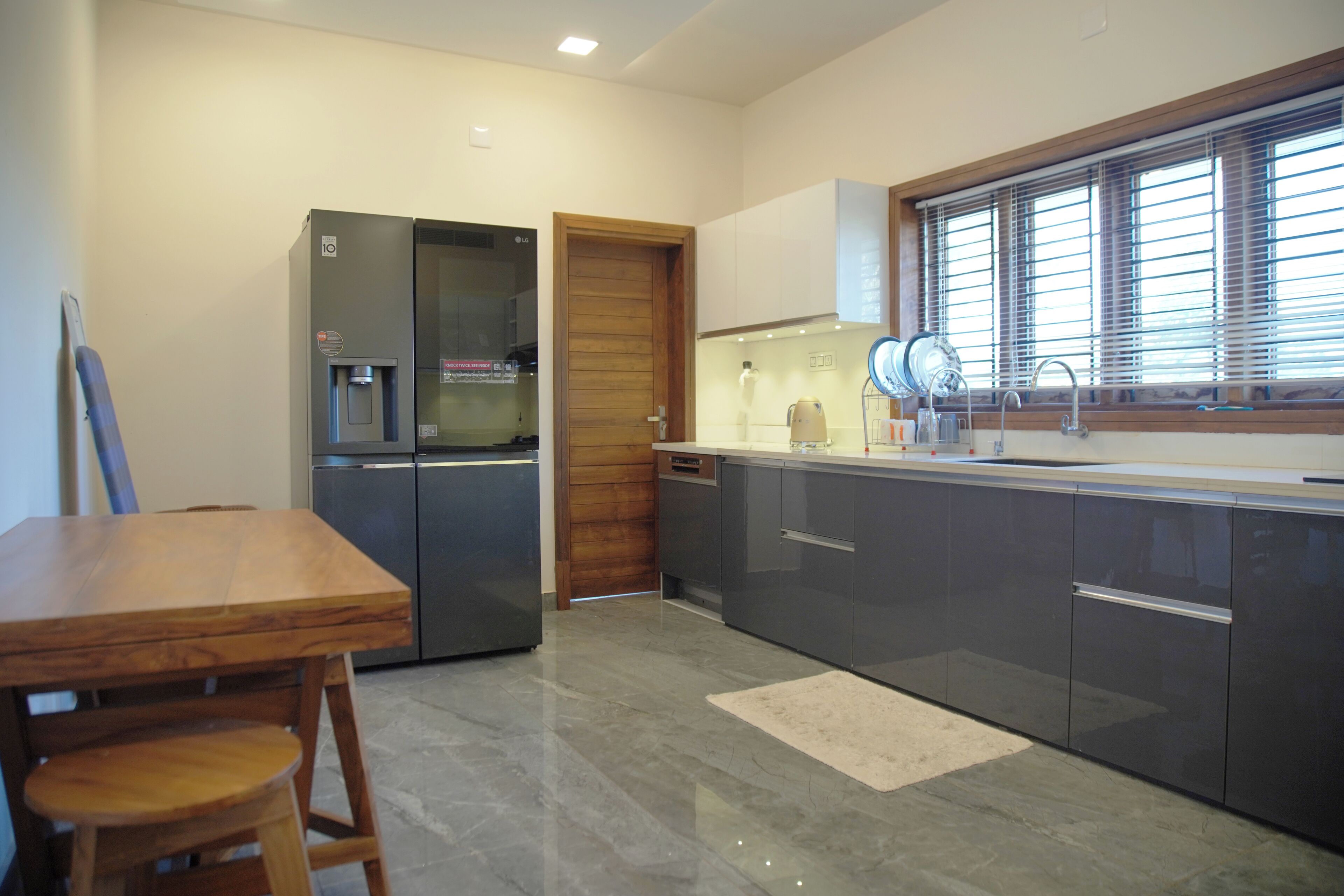 Uma cozinha moderna com uma mesa de jantar e cadeiras de madeira, uma geladeira embutida e uma janela com persianas.