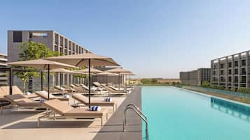 Uma área de piscina moderna com espreguiçadeiras e guarda-sóis, piscina azul límpida e edifícios contemporâneos ao fundo.