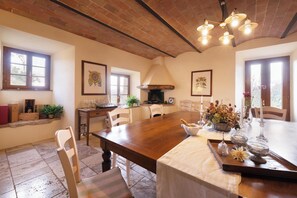 Uma sala de jantar com uma mesa de madeira, cadeiras e um candelabro. Há quadros nas paredes e uma janela com vista para o verde.