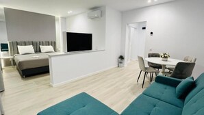 Uma sala de estar moderna com um sofá, uma mesa de jantar com cadeiras, uma TV fixada na parede e uma pequena planta.