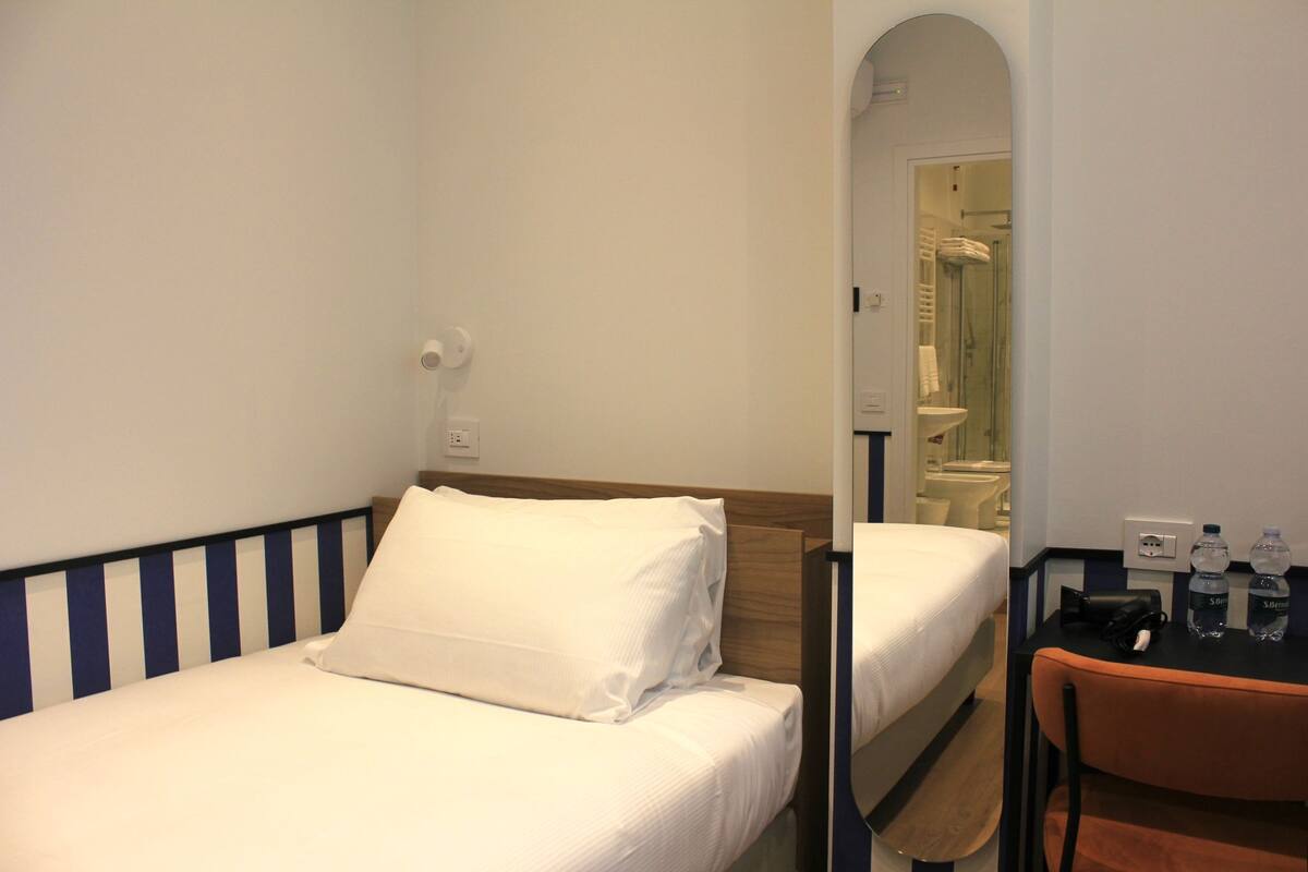 Quarto de hotel com duas camas de solteiro, uma mesa pequena com uma cadeira e um espelho refletindo o banheiro.