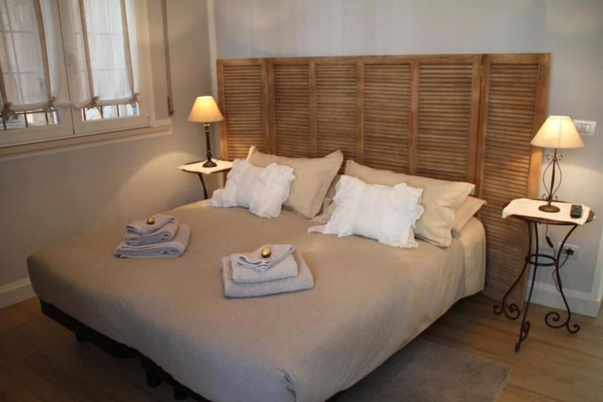 Uma cama bem arrumada com cabeceira de madeira, duas mesas de cabeceira com abajures e toalhas dobradas sobre a cama.