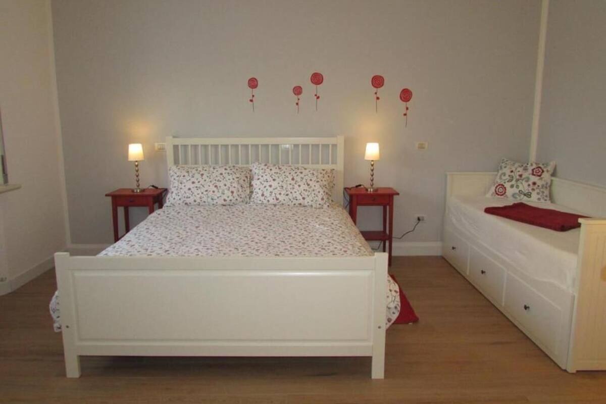 Quarto com cama branca, duas mesinhas de cabeceira, um travesseiro vermelho e parede com detalhes vermelhos.