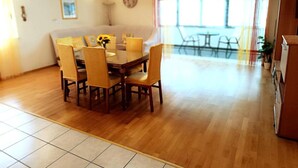 Uma área de refeições com mesa e cadeiras de madeira, um relógio na parede e vista para o exterior por grandes janelas.